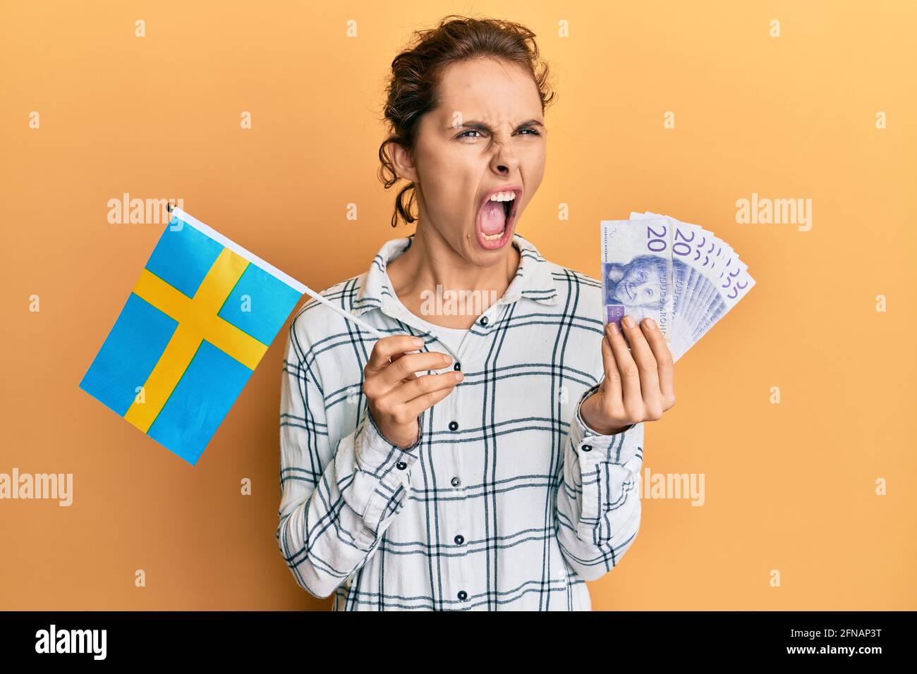 Young brunette woman holding sweden flag and krone banknotes angry and ...