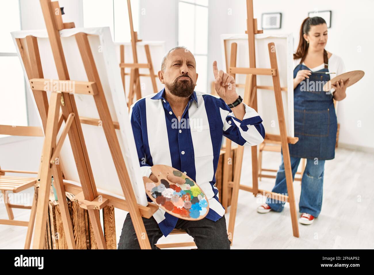 Senior artist man at art studio pointing up looking sad and upset ...