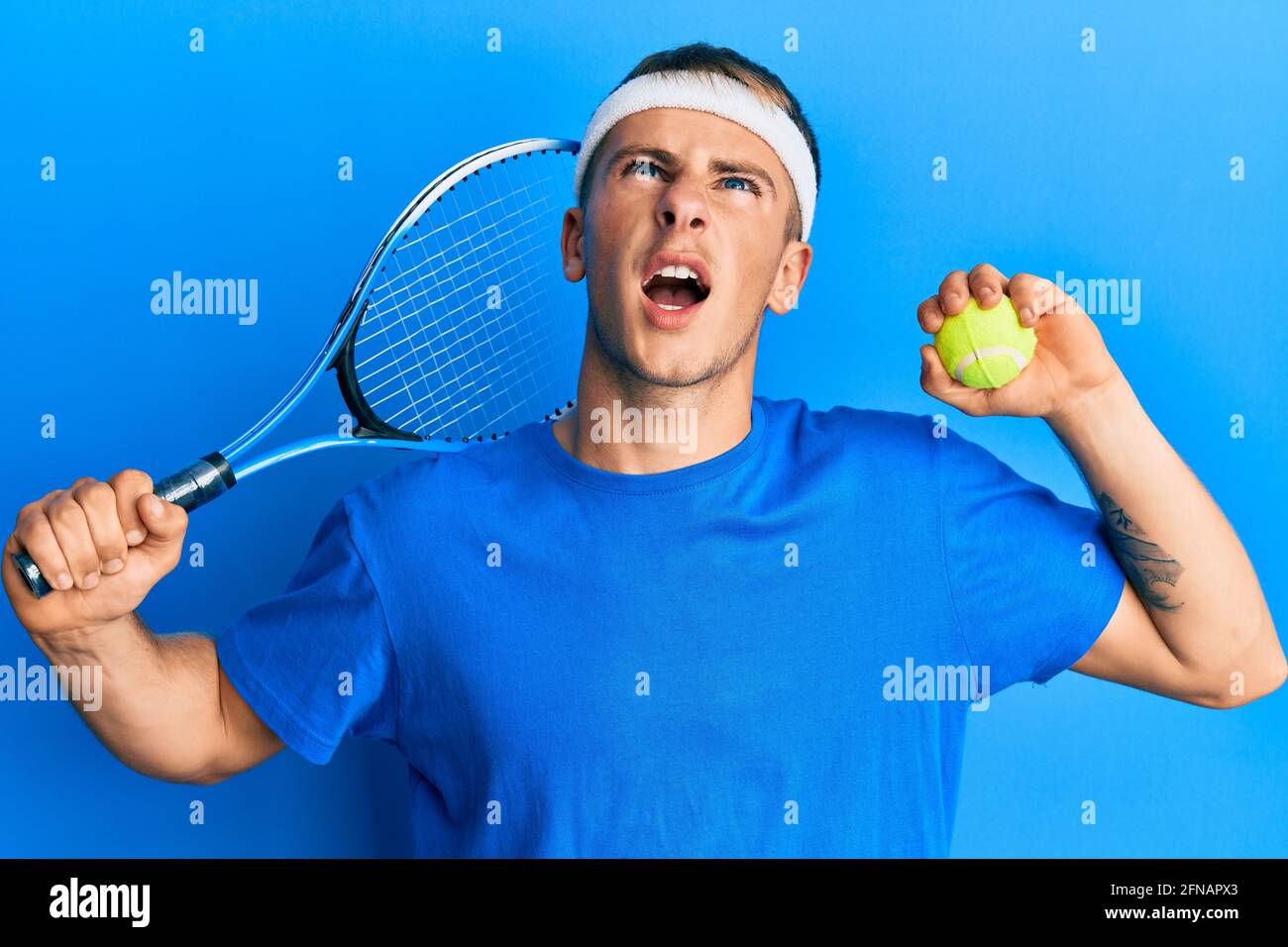 Young caucasian man playing tennis holding racket and ball angry and ...