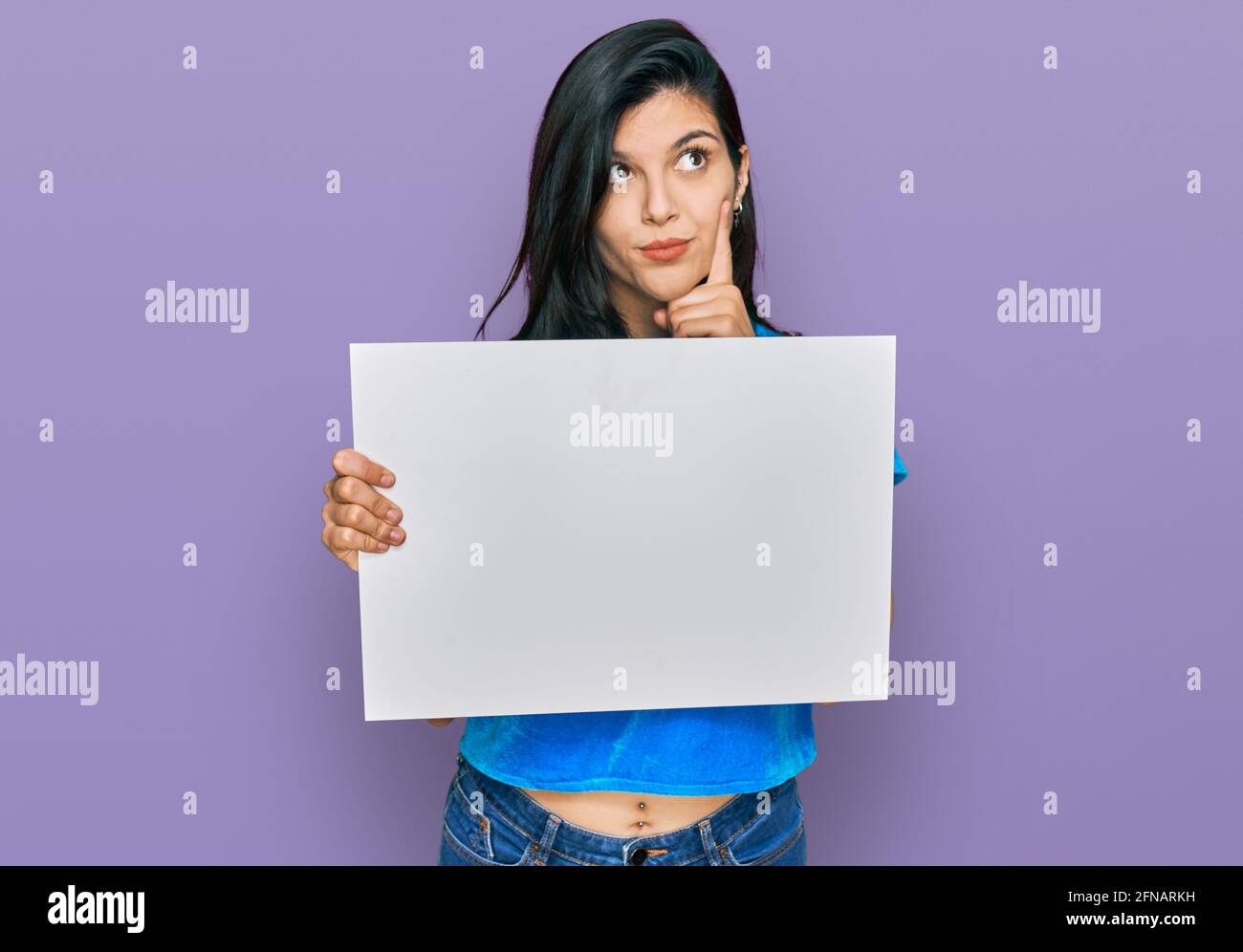 Young hispanic woman holding blank empty banner serious face thinking ...