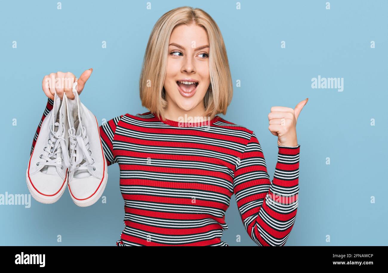 Young blonde woman holding white casual shoes pointing thumb up to the ...