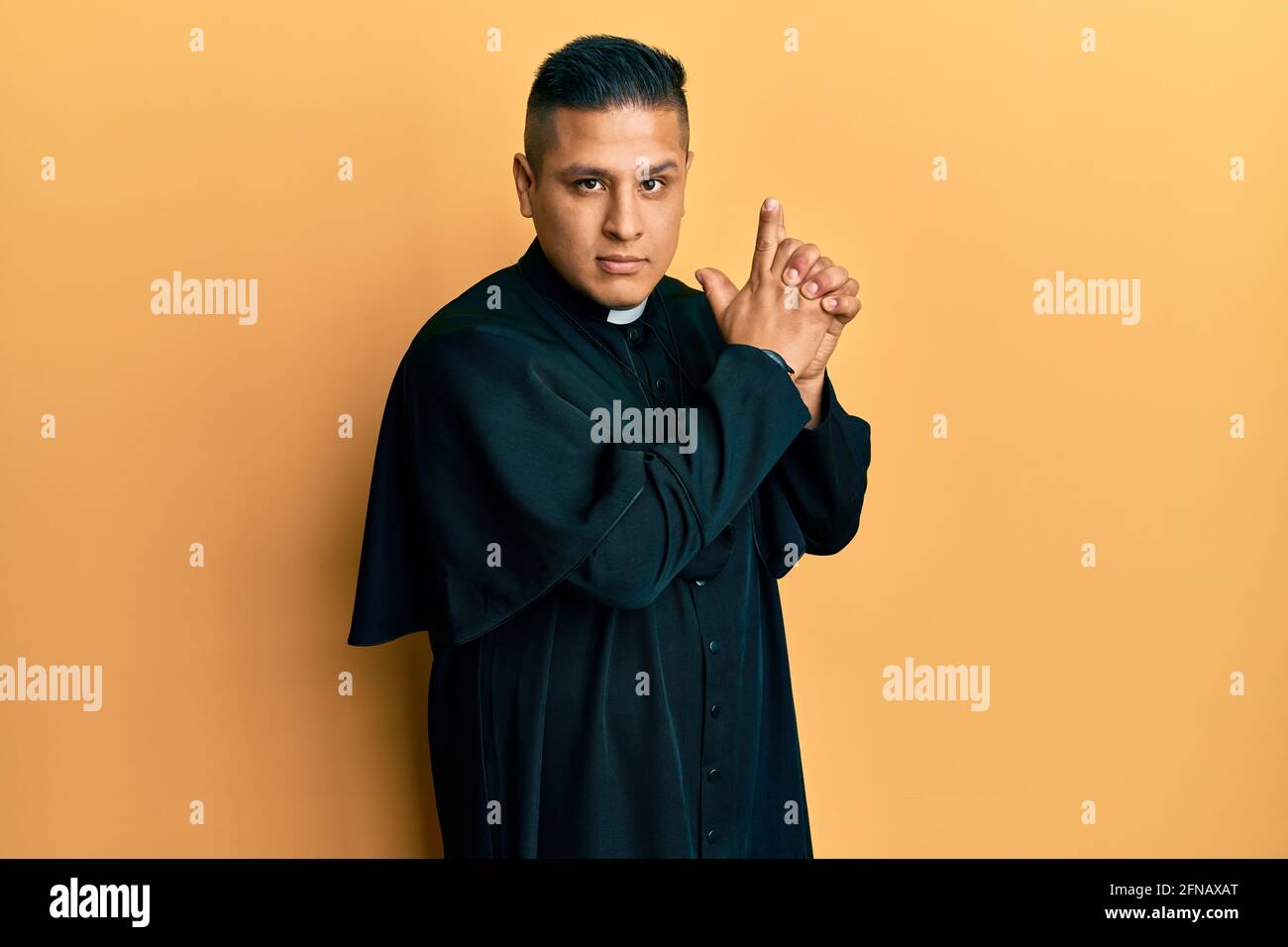 Young latin priest man standing over yellow background holding symbolic ...