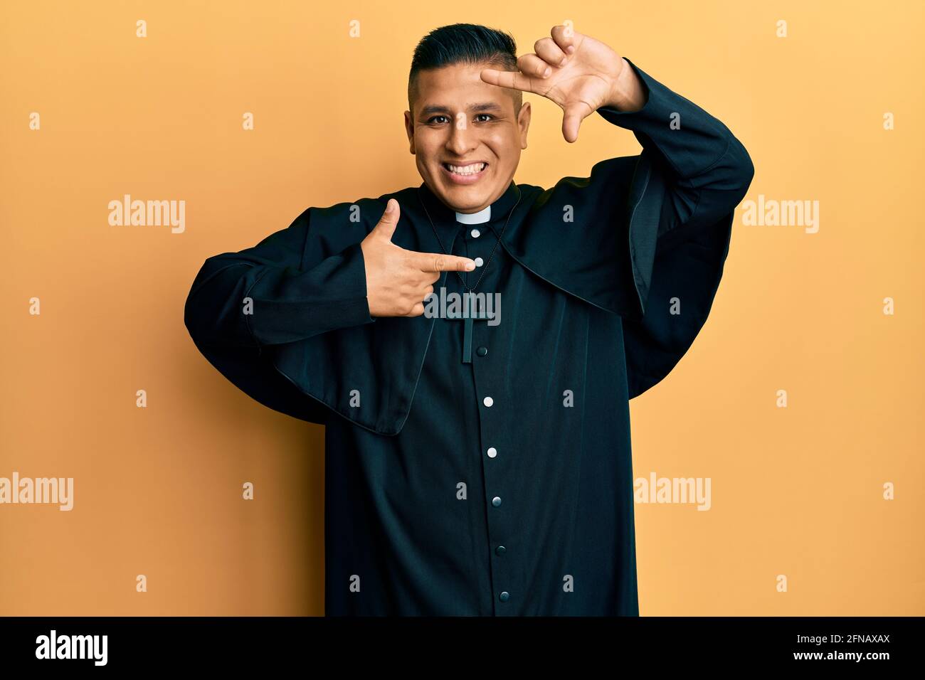 Young latin priest man standing over yellow background smiling making ...