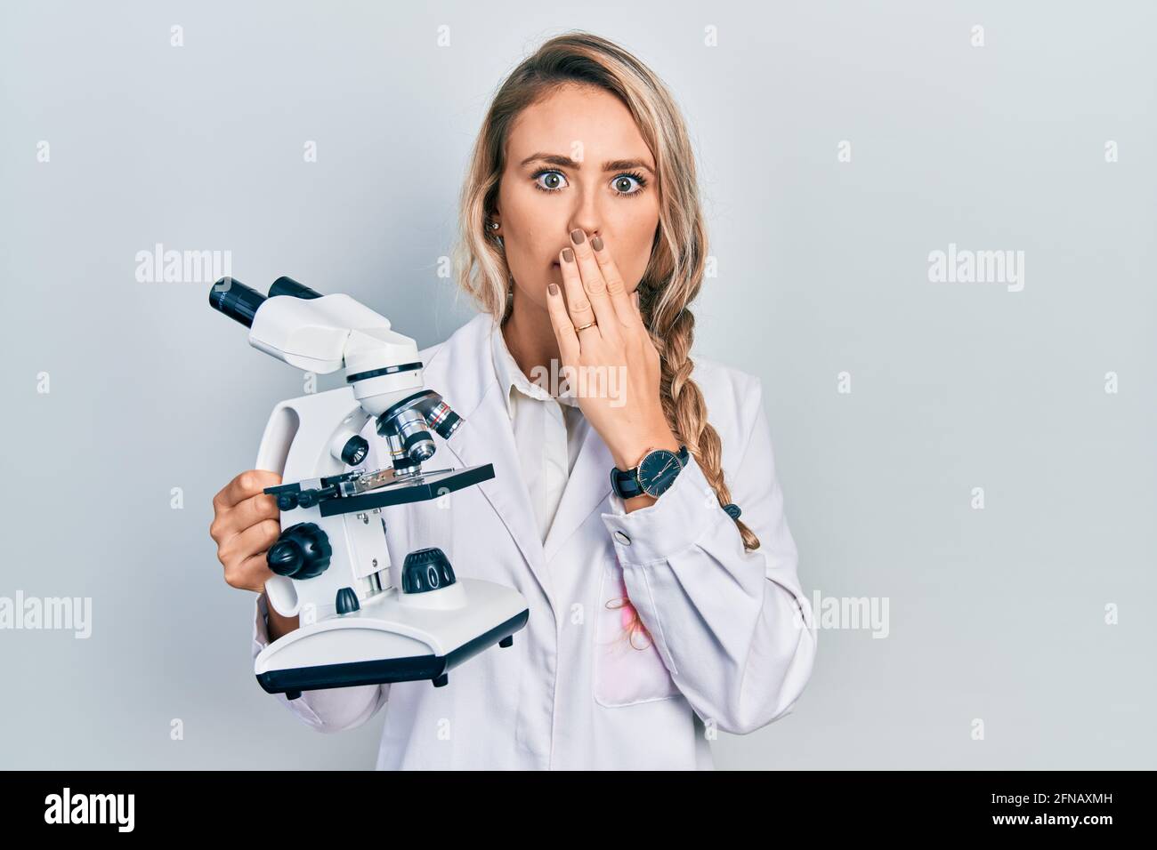 Beautiful young blonde woman holding microscope covering mouth with ...