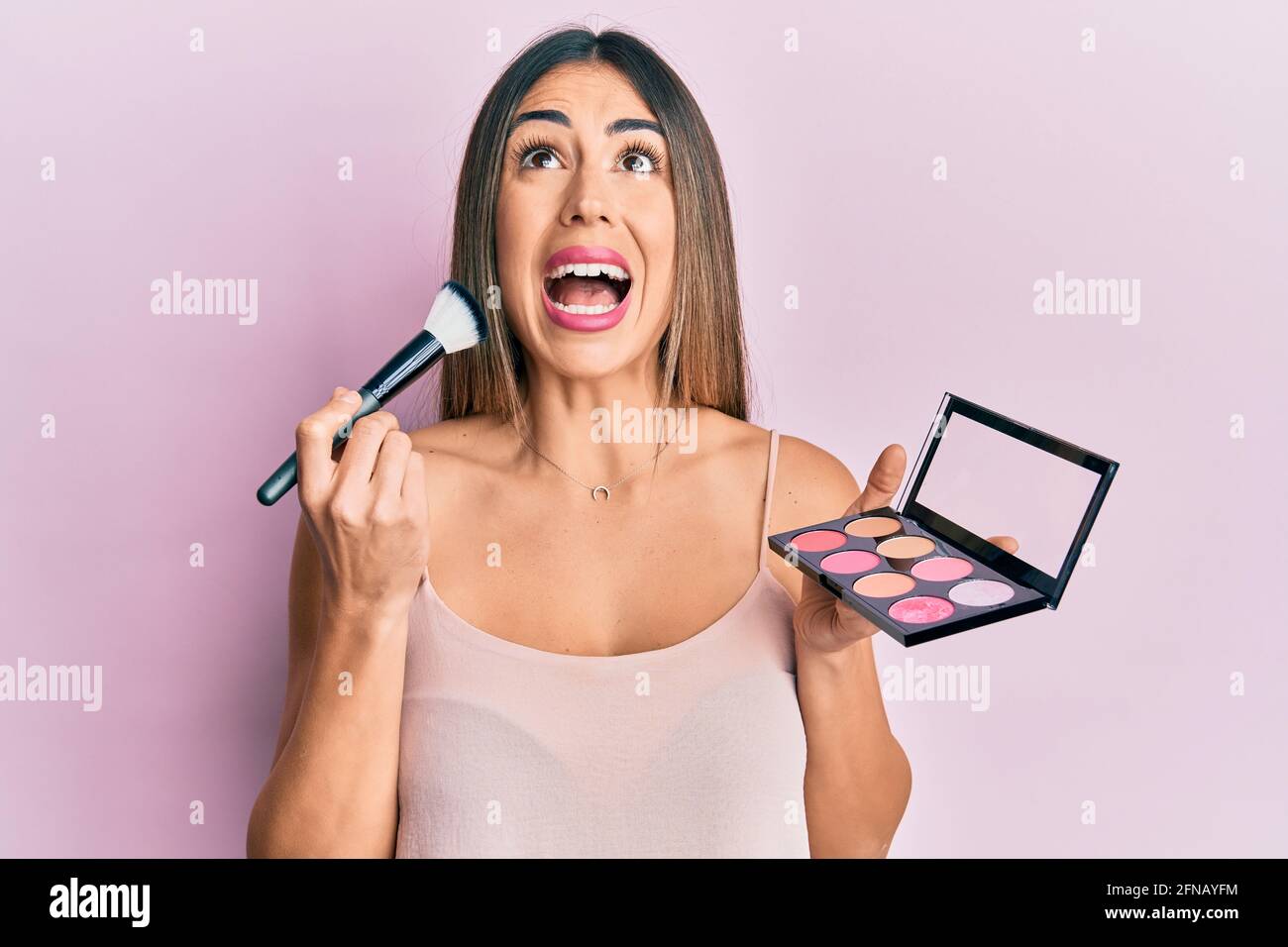Young hispanic woman holding makeup brush and blush angry and mad ...