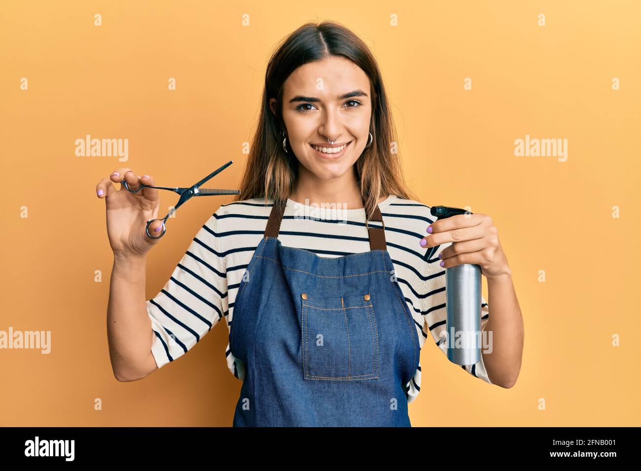 Young hispanic woman wearing hairdresser apron and holding scissors smiling with a happy and ...