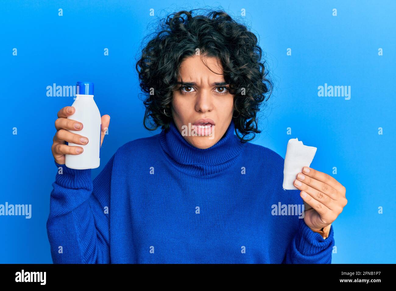 Young hispanic woman with curly hair holding medical alcohol in shock ...