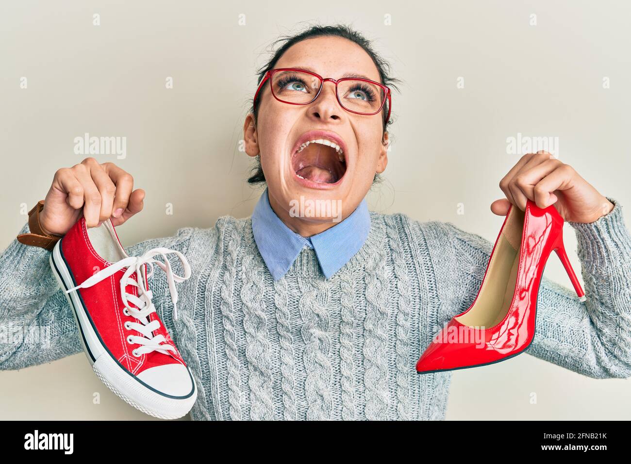 Young caucasian woman choosing high heel shoes and sneakers angry and mad screaming frustrated ...