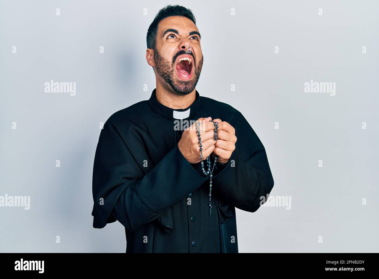 Handsome hispanic priest man with beard praying holding catholic rosary ...