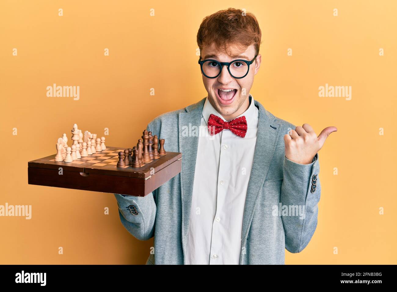 Young caucasian man wearing glasses holding chess board pointing thumb ...