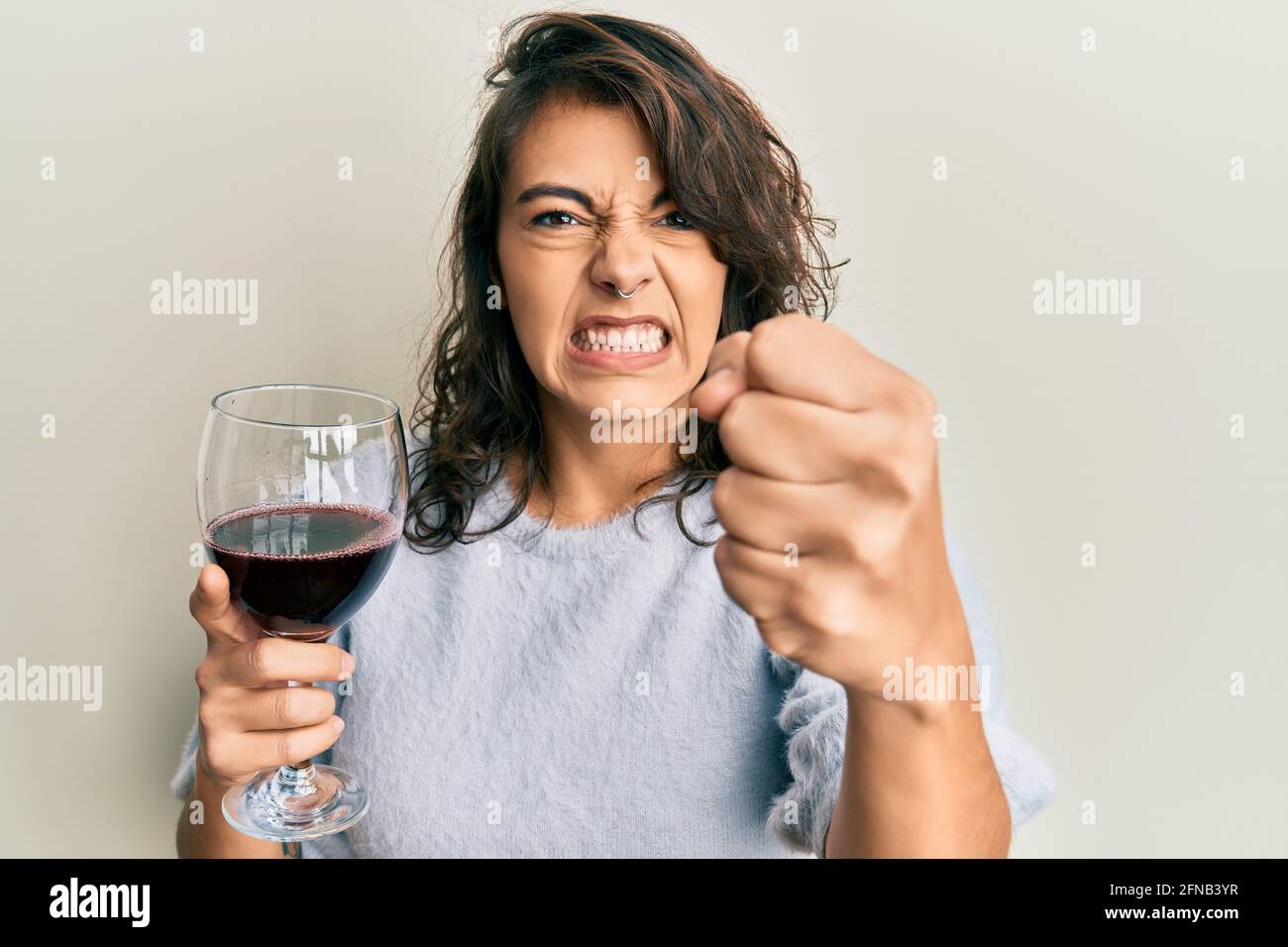 Young hispanic woman drinking a glass of red wine annoyed and ...