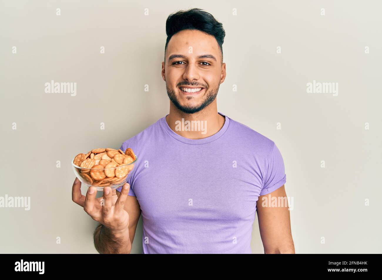 Young arab man holding bowl of salty crackers biscuits looking positive ...