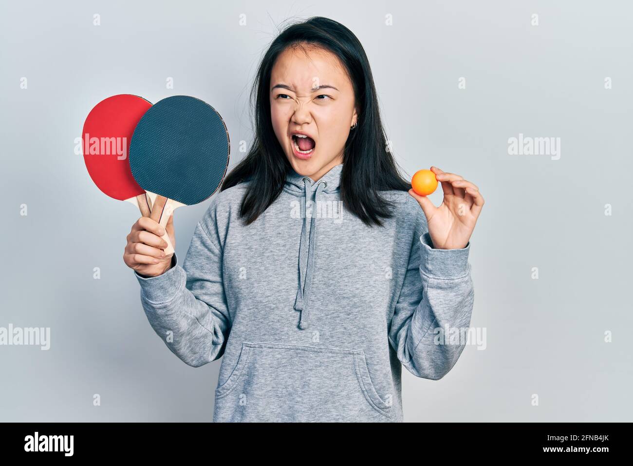 Young chinese girl holding red ping pong rackets and ball angry and mad ...