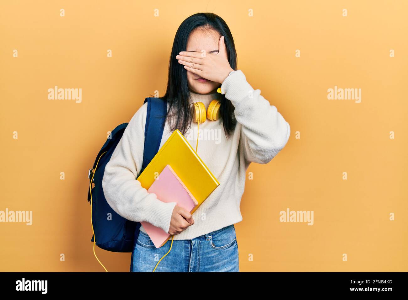 Young chinese girl holding student backpack and books covering eyes ...