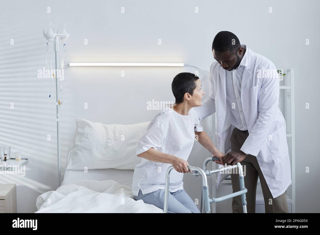 African young doctor helping his patient to stand up at hospital ward ...