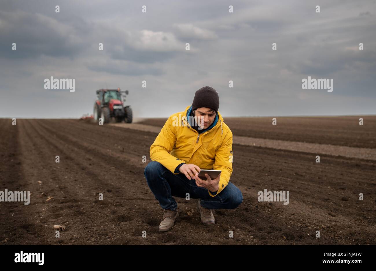Portrait of handsome farmer with tablet squatting in front of tractor ...
