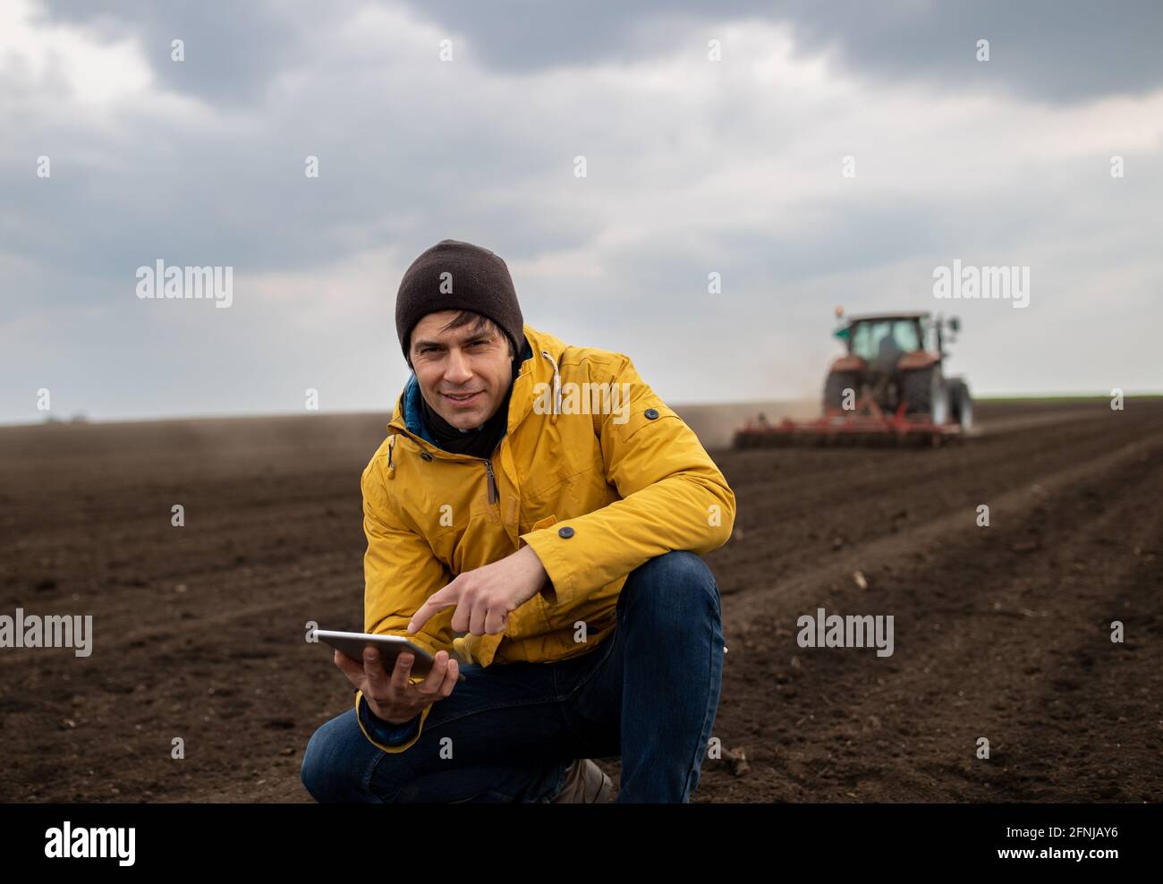 Portrait of handsome farmer with tablet squatting in front of tractor ...