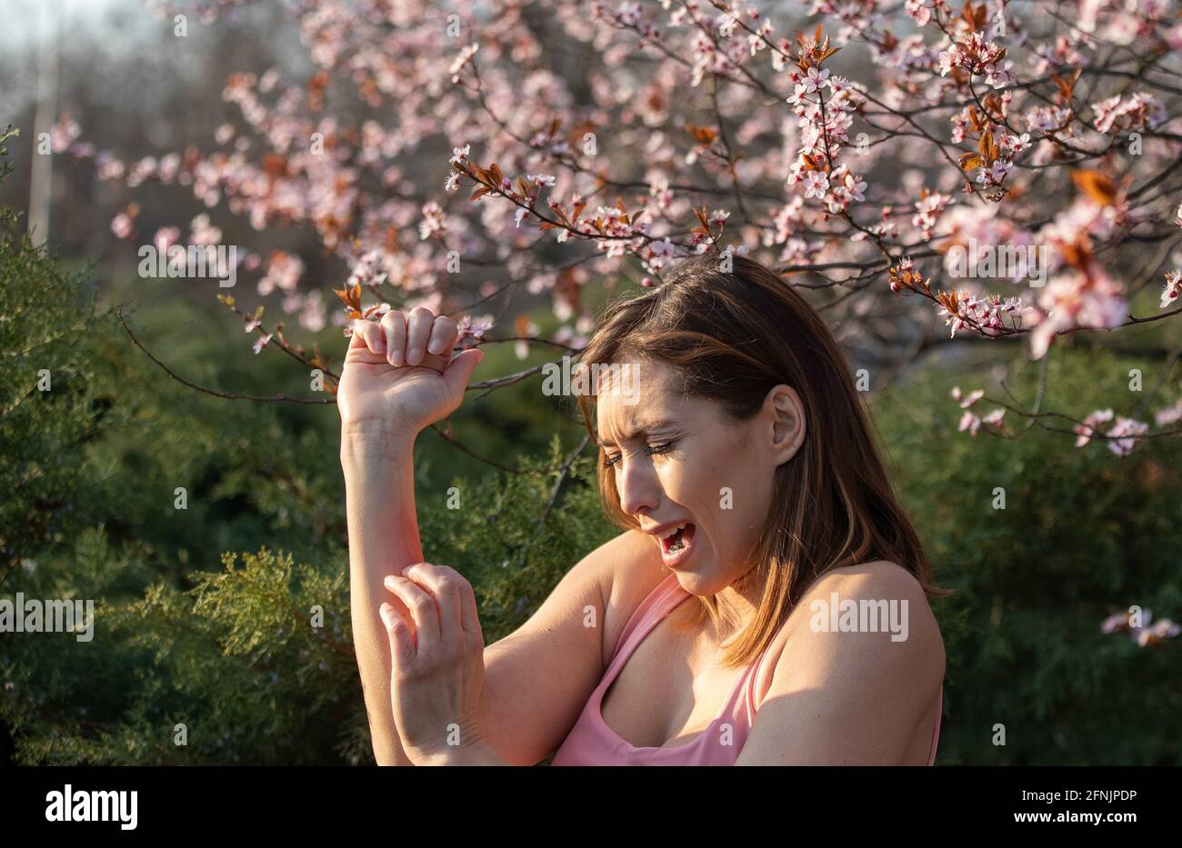 Pretty young woman scratching arm beside blooming tree in park in ...