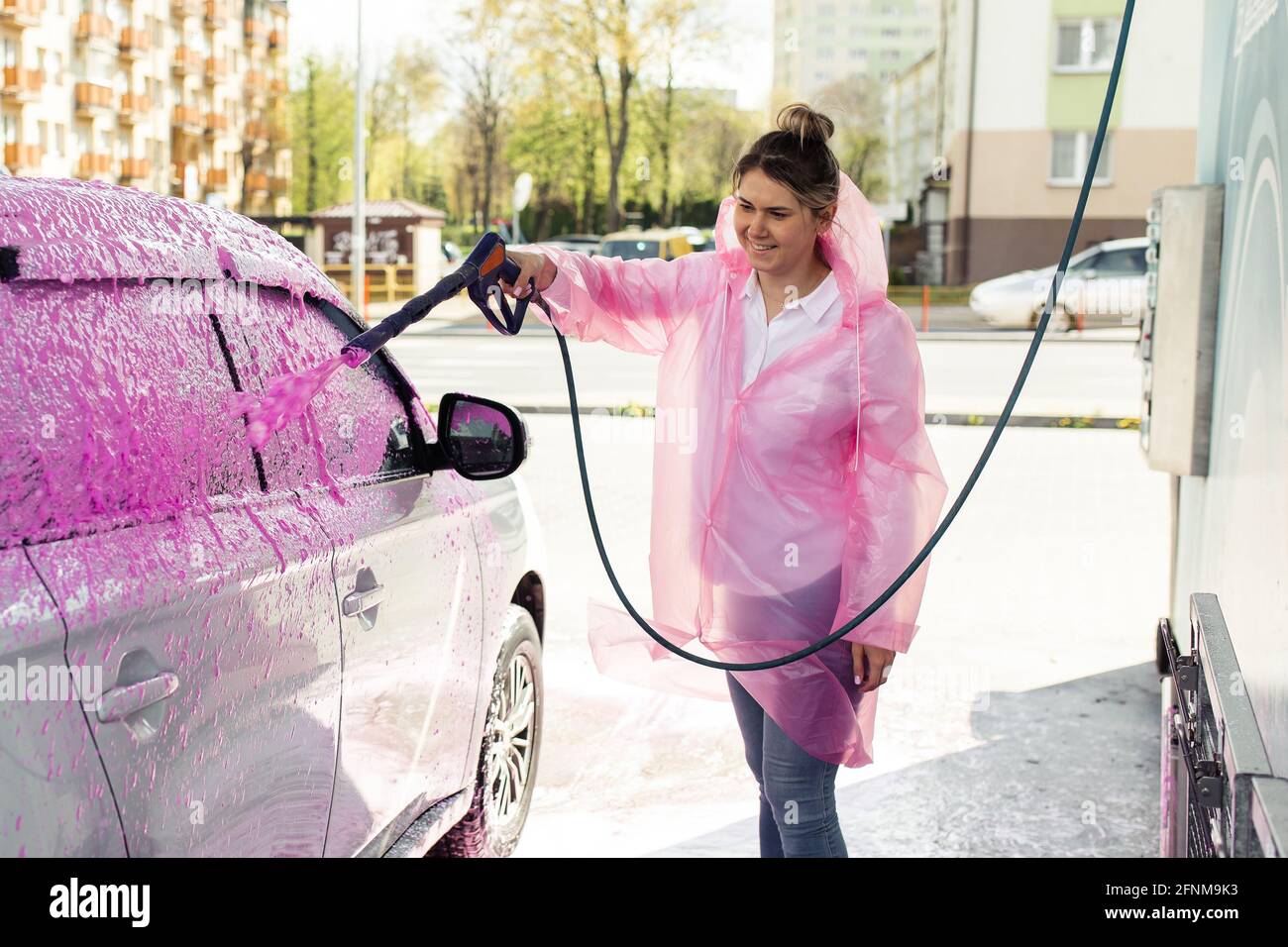 Young woman in a pink raincoat washes car at self-service car wash ...