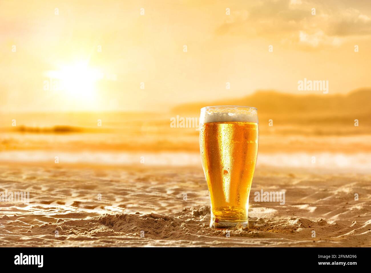 Glass with fresh beer on the sand on the beach with golden sunset ...