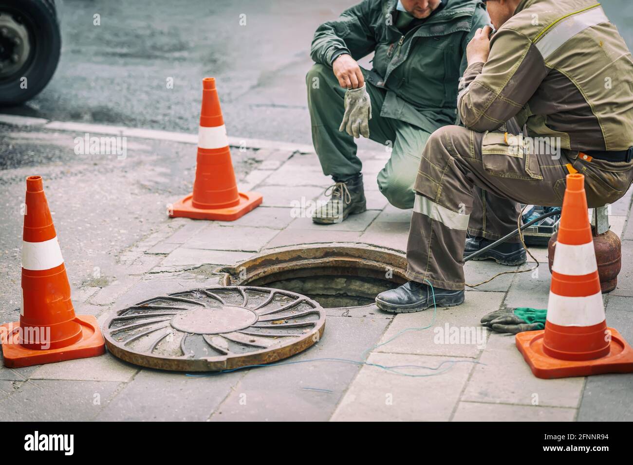 Workers sitting on over open sewer hatch on street near traffic cones ...