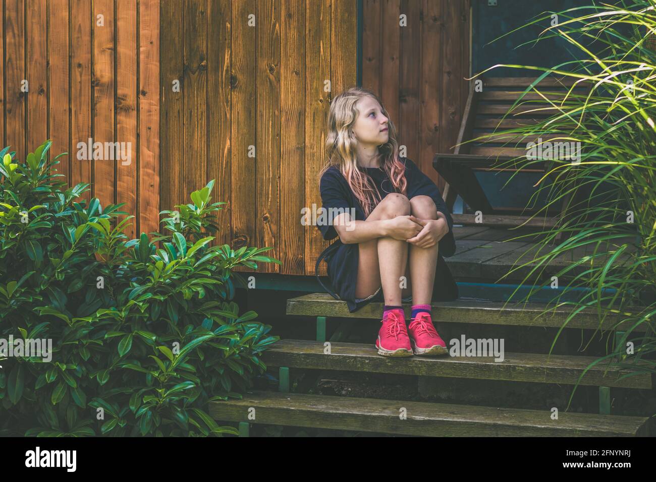 adorable blond girl sitting on stairs by wooden cottage in green garden ...
