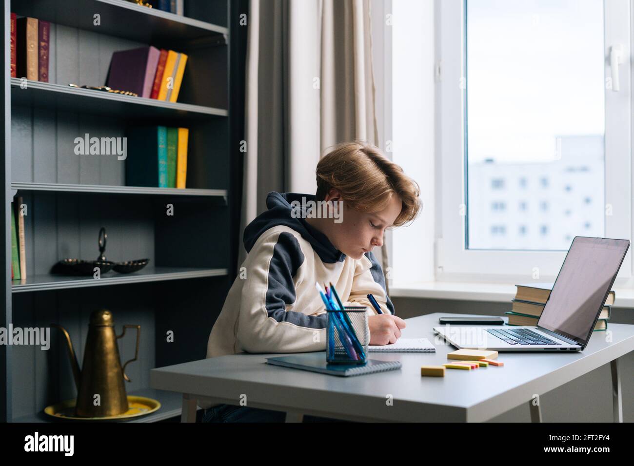 Side view of pupil boy making notes in copybook with pen sitting at ...