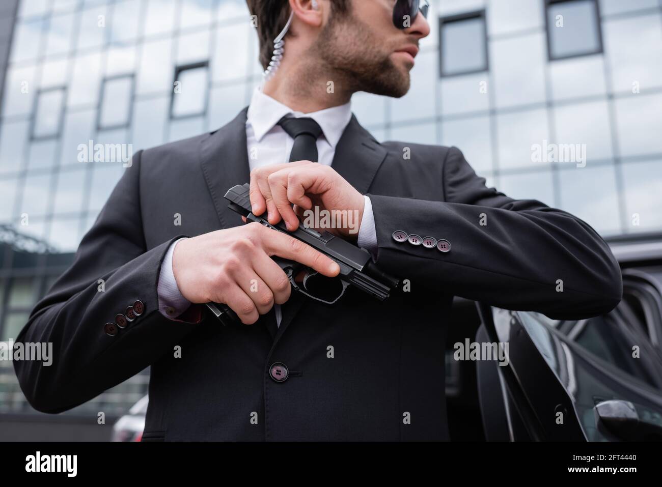 side view of bearded bodyguard in suit holding gun near modern building ...