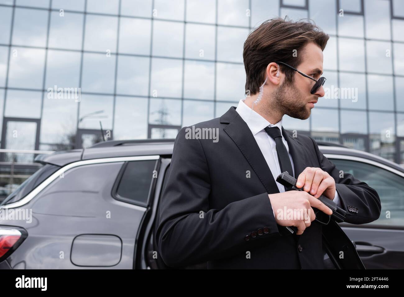 bearded bodyguard in sunglasses and suit holding gun near modern ...