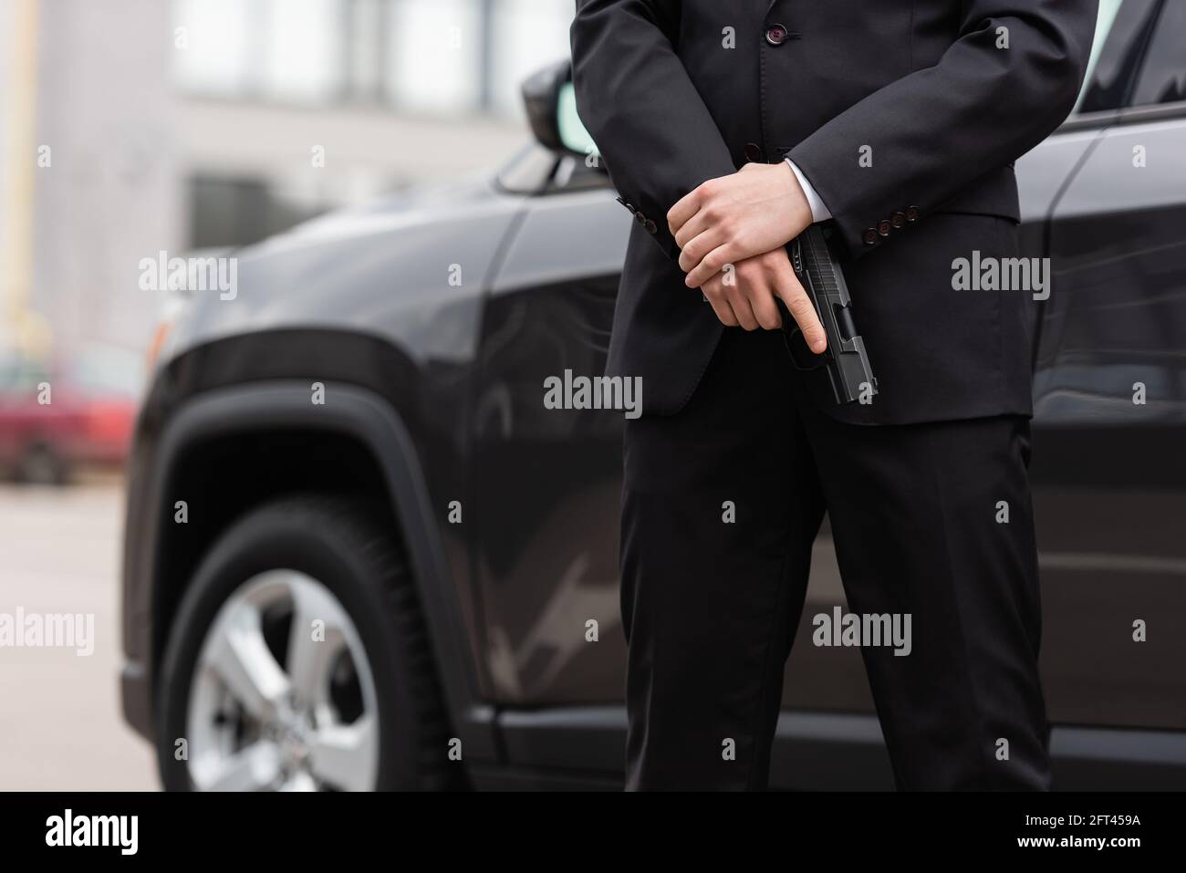 cropped view of bodyguard in suit holding gun near blurred modern auto ...