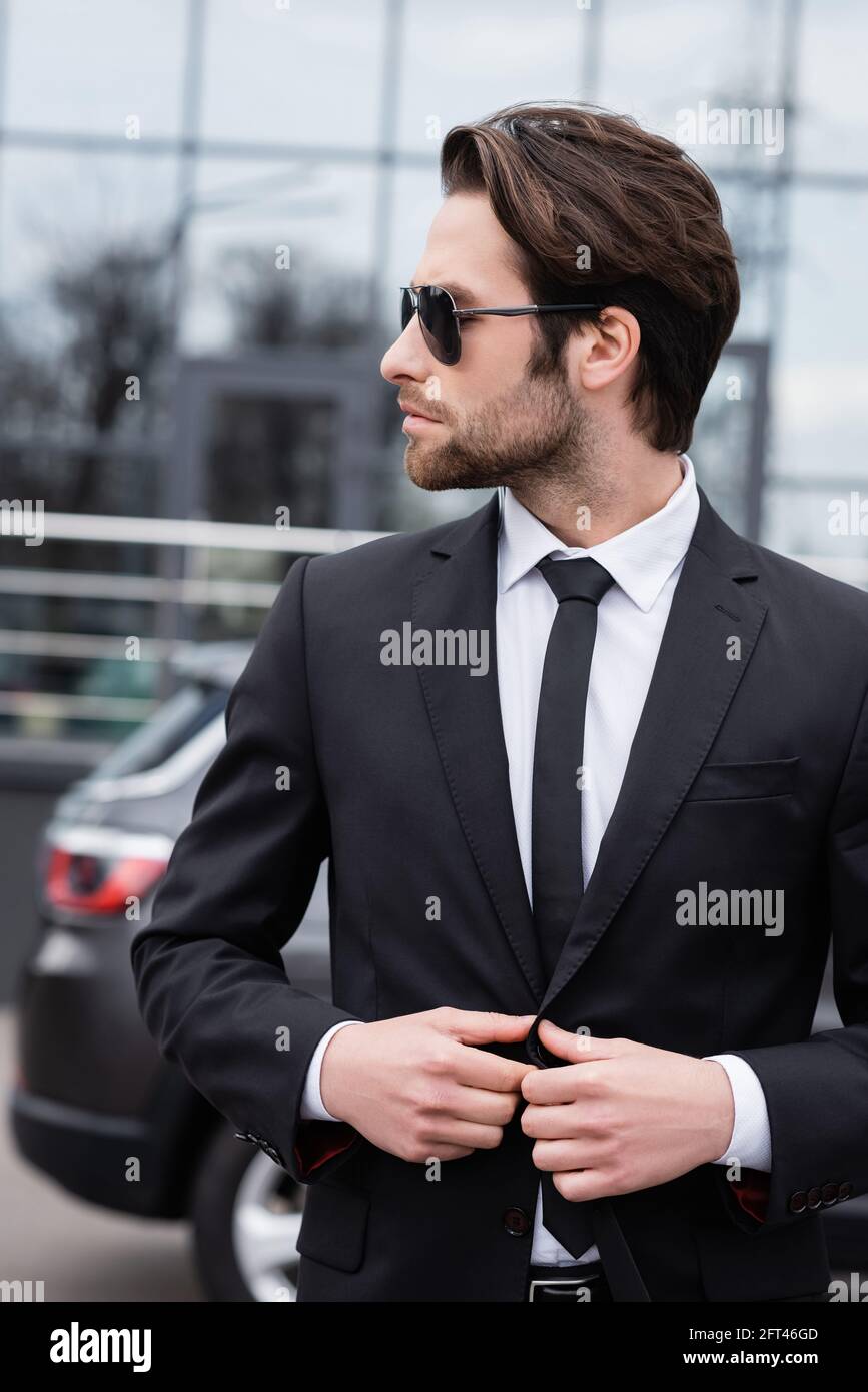 side view of bearded safeguard in suit and sunglasses near modern car ...