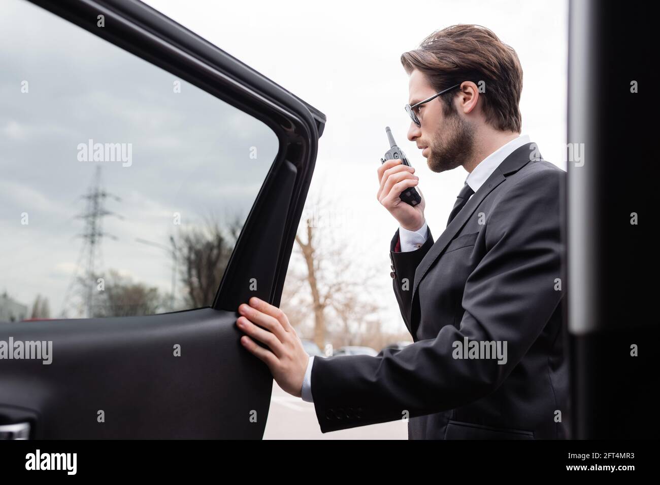 side view of bearded bodyguard in suit and sunglasses using walkie ...