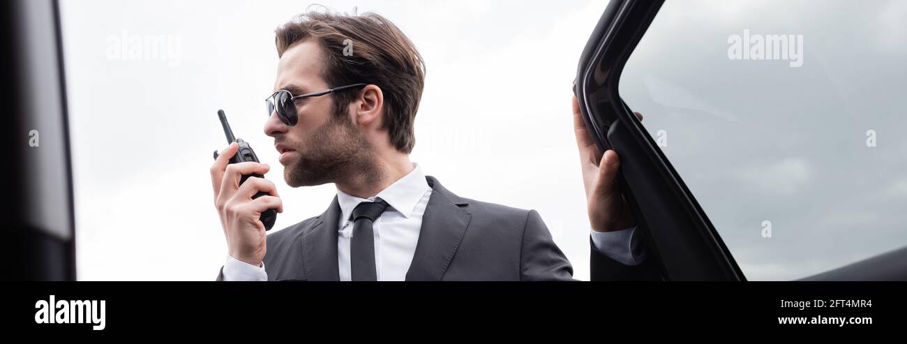 side view of bearded bodyguard in suit and sunglasses using walkie ...