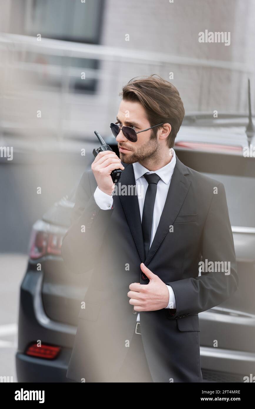 bodyguard in sunglasses and suit using walkie talkie near blurred ...
