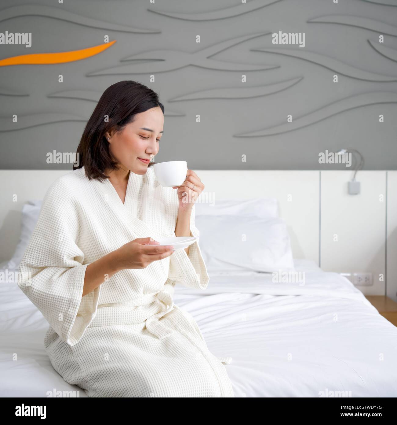 Asian woman in a bathrobe drinking hot coffee on bed in the morning ...