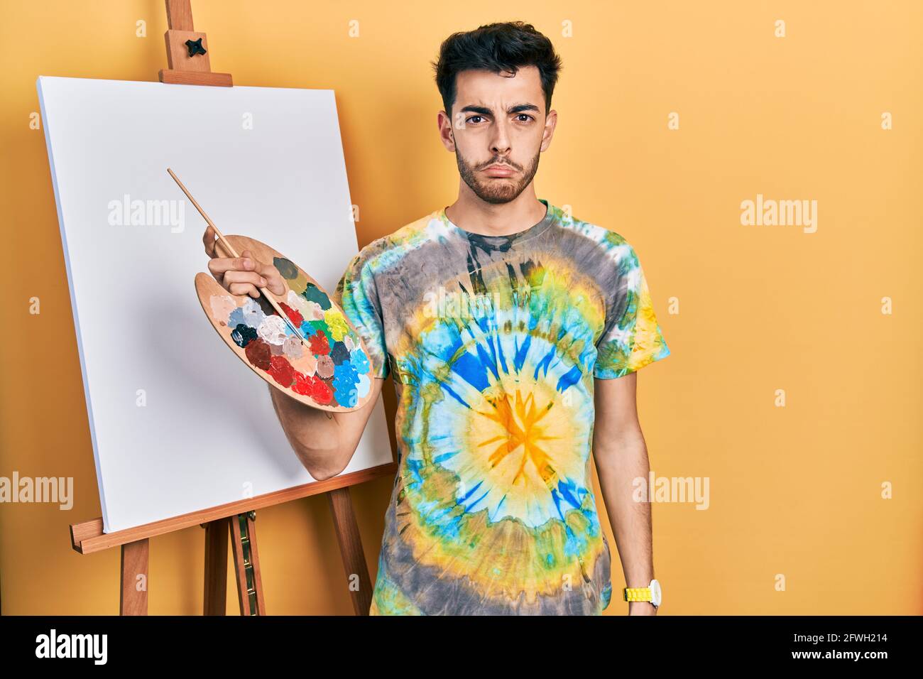 Young hispanic man standing drawing with palette by painter easel stand ...