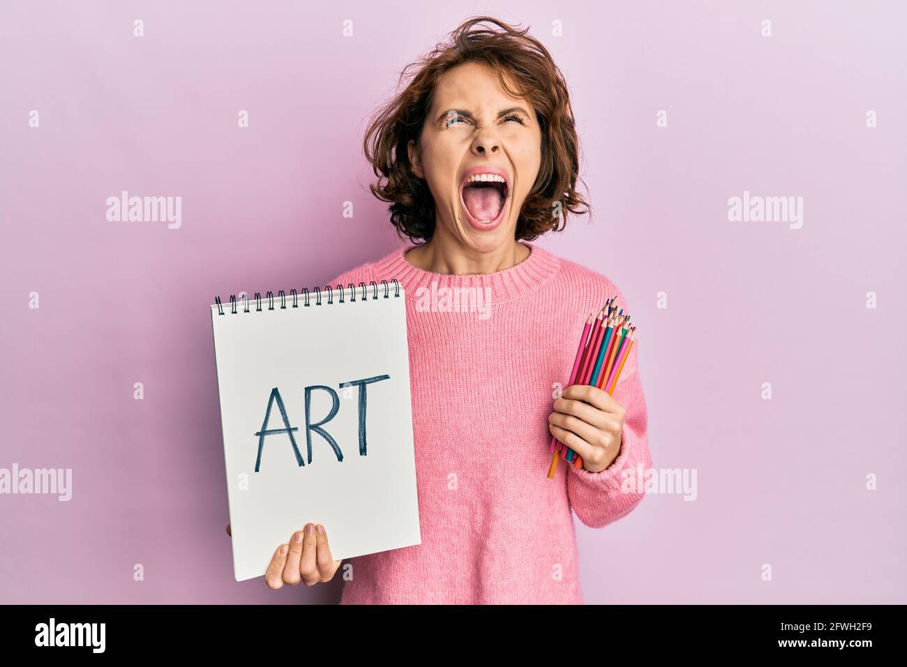 Young brunette woman holding art notebook and colored pencils angry and ...