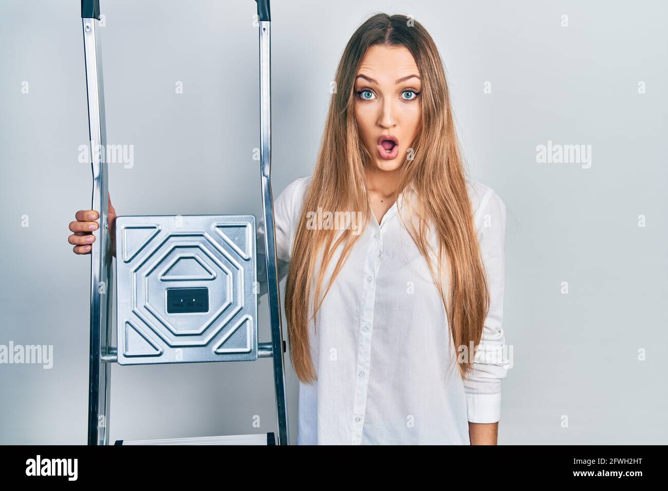 Young blonde girl holding ladder scared and amazed with open mouth for surprise, disbelief face ...