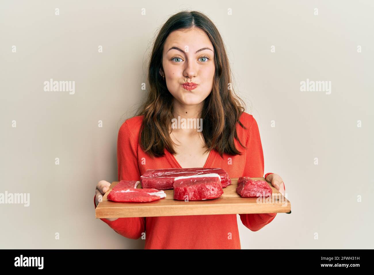 Young brunette woman holding board with raw meat puffing cheeks with ...