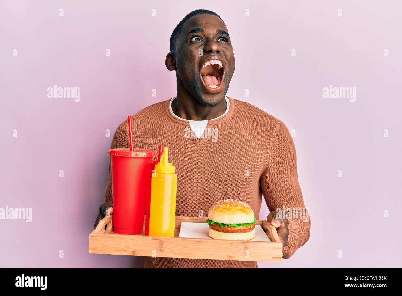 Young african american man eating a tasty classic burger and soda angry ...