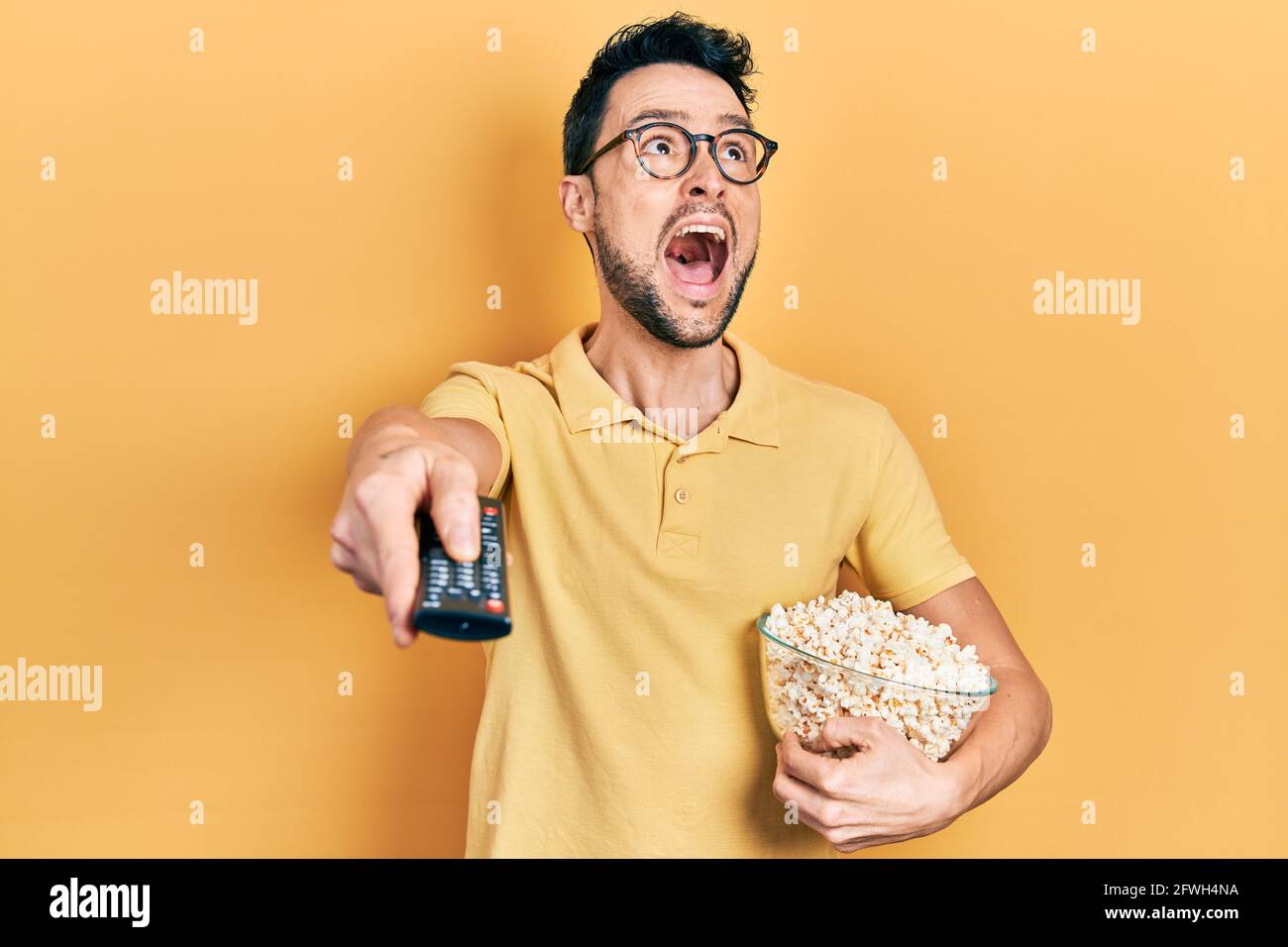 Young hispanic man eating popcorn using tv control angry and mad ...