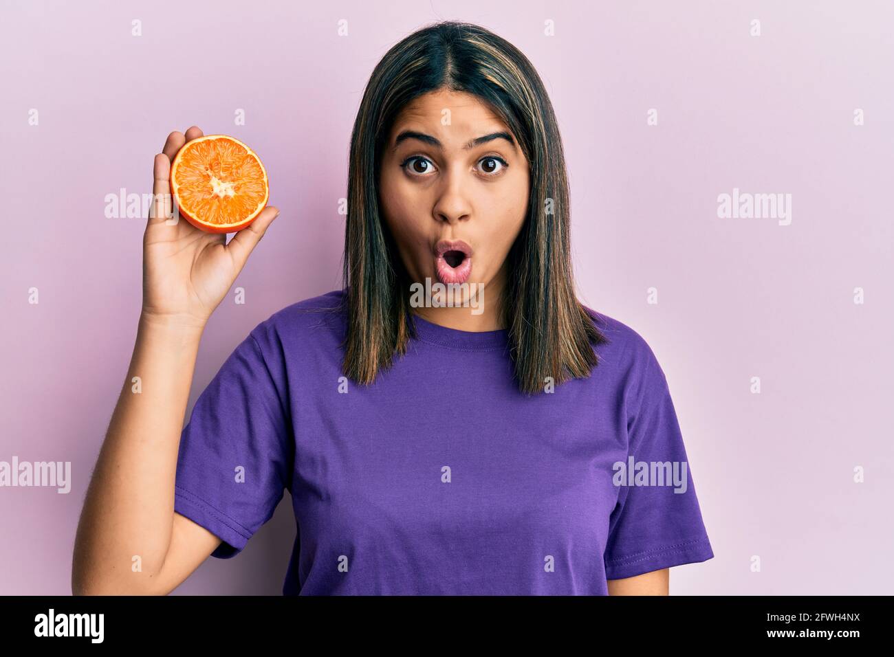 Young latin woman holding fresh orange slice scared and amazed with ...