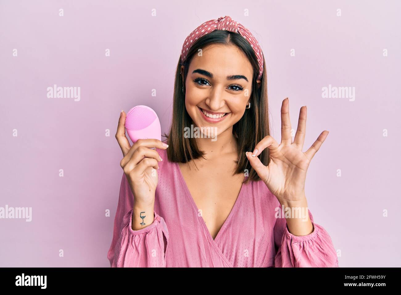 Young brunette girl using facial exfoliating cleaner doing ok sign with ...