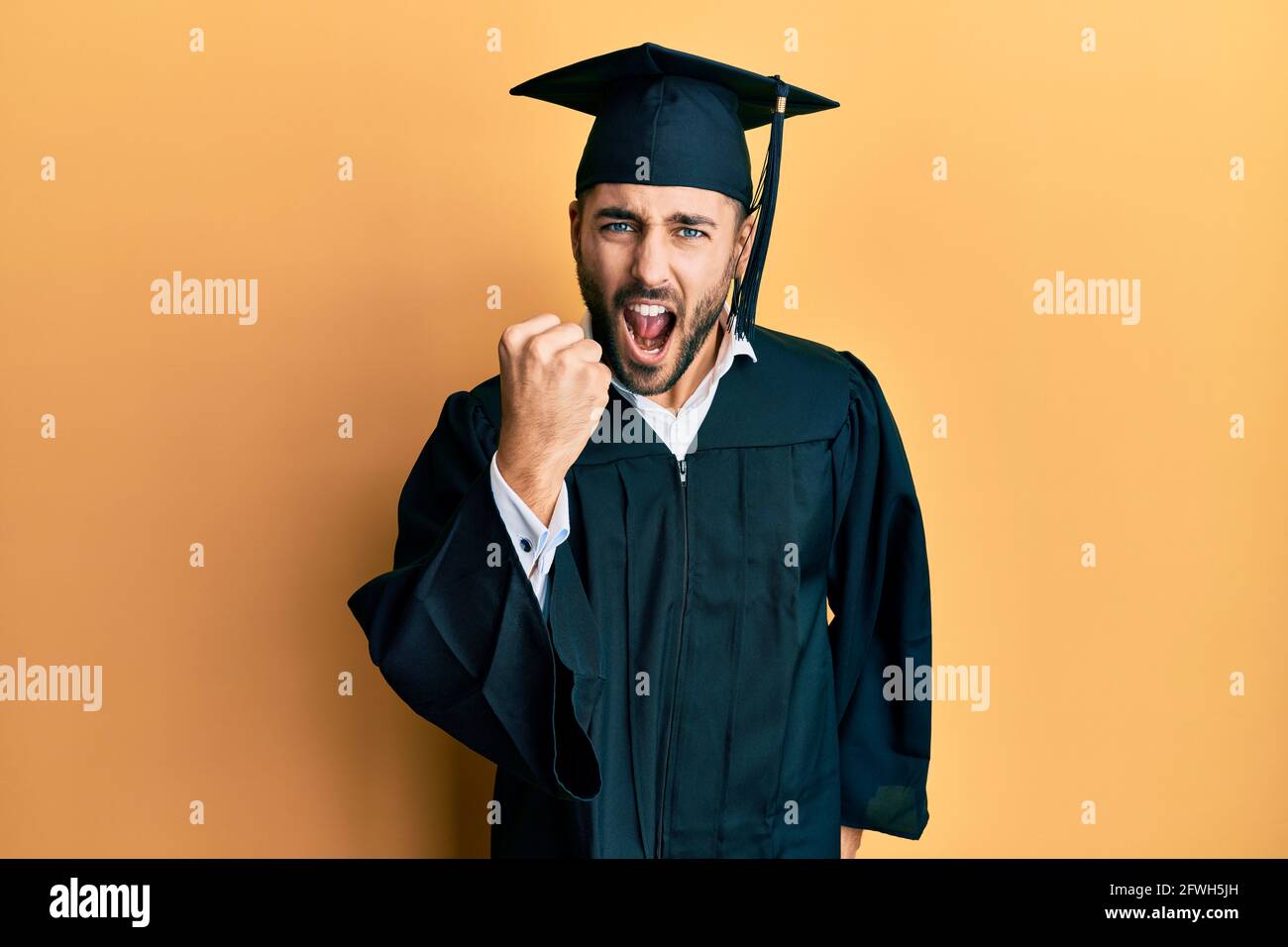 Young hispanic man wearing graduation cap and ceremony robe angry and ...