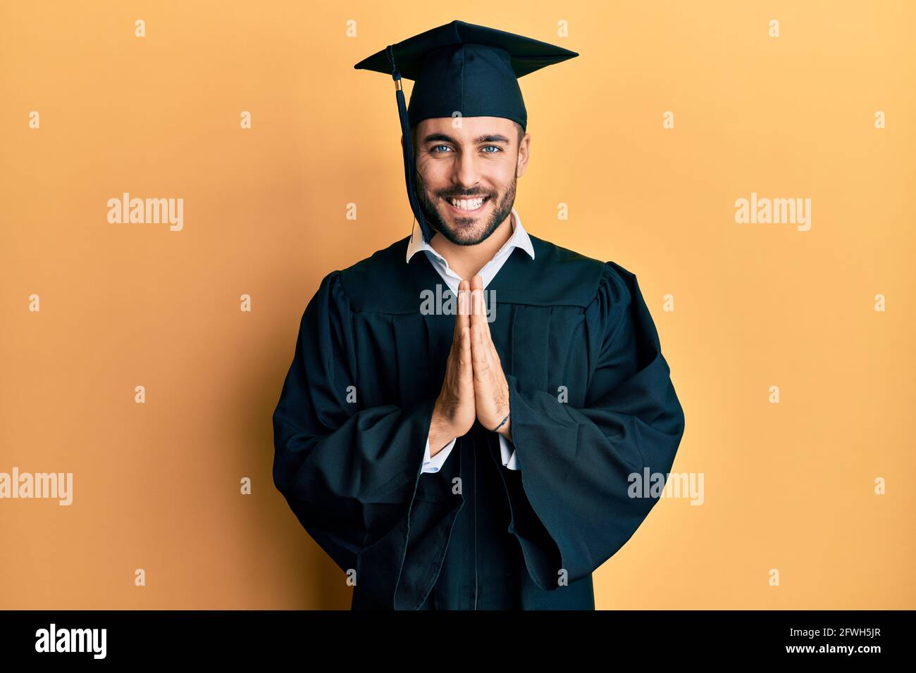 Young hispanic man wearing graduation cap and ceremony robe praying ...