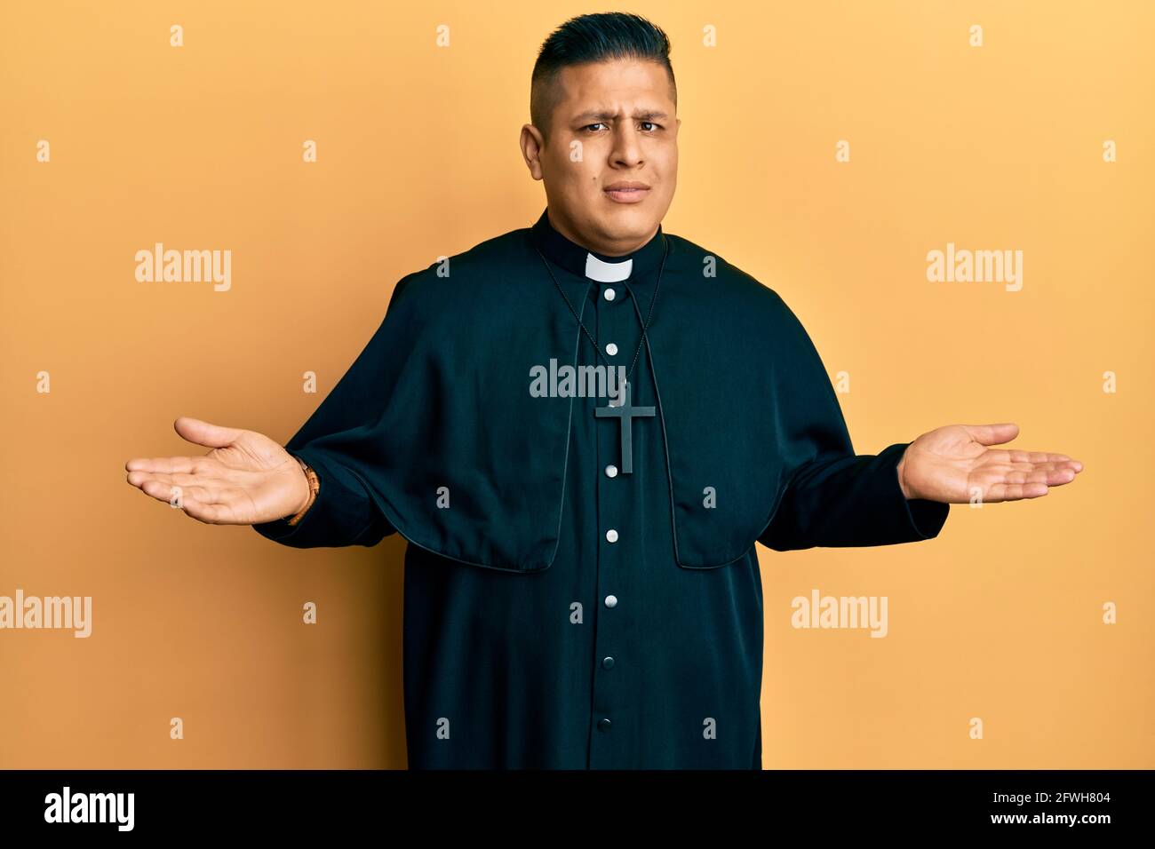 Young latin priest man standing over yellow background clueless and ...