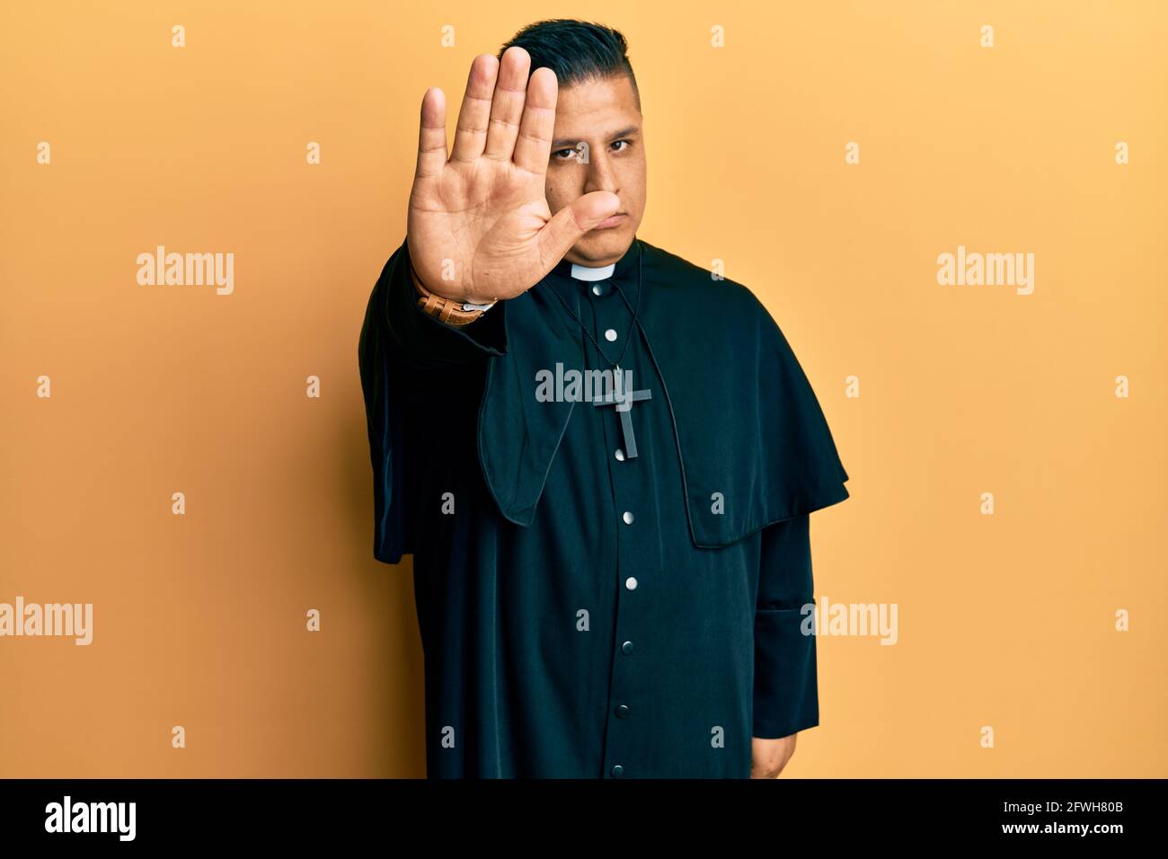 Young latin priest man standing over yellow background doing stop sing ...