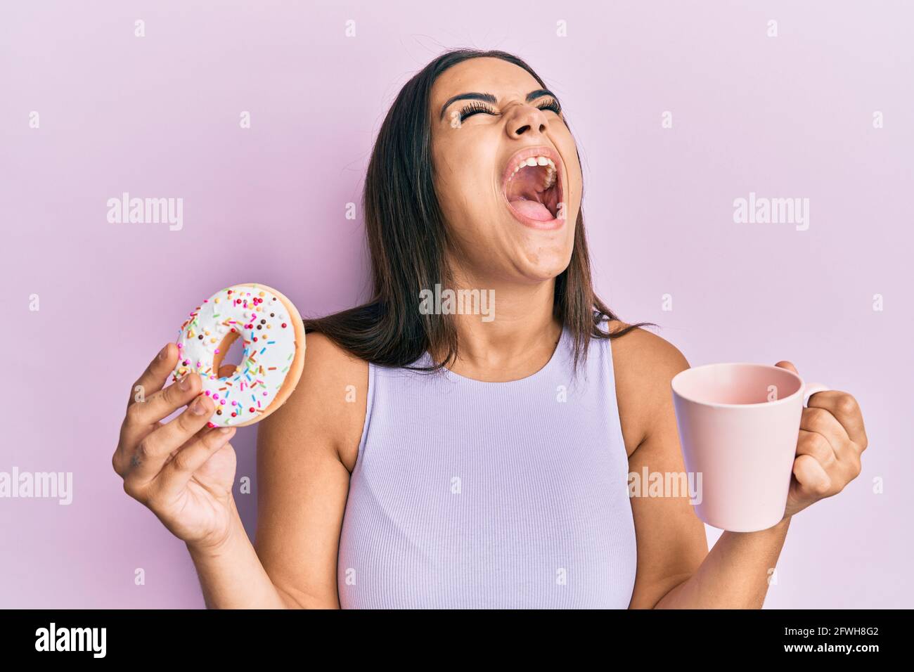 Young brunette woman eating doughnut and drinking coffee angry and mad ...