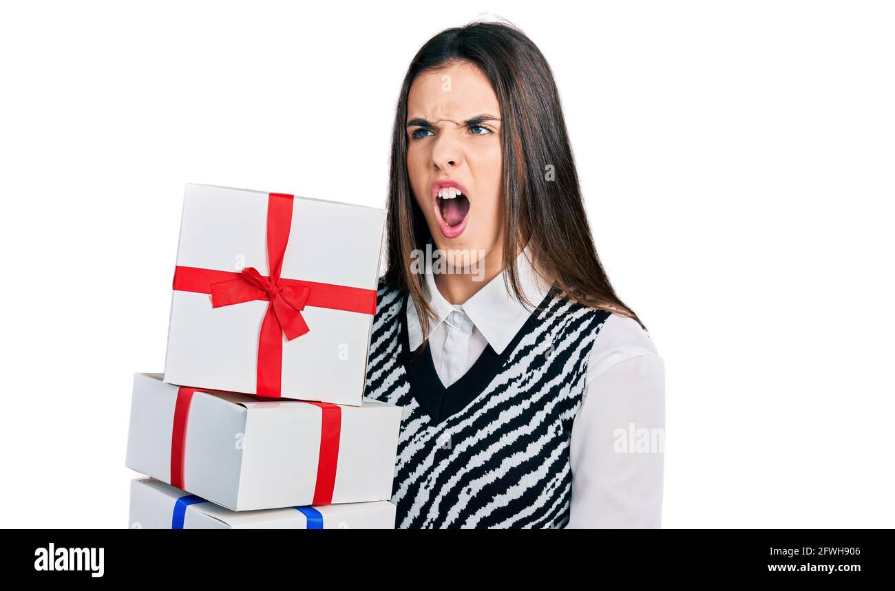 Young brunette teenager holding gifts angry and mad screaming ...