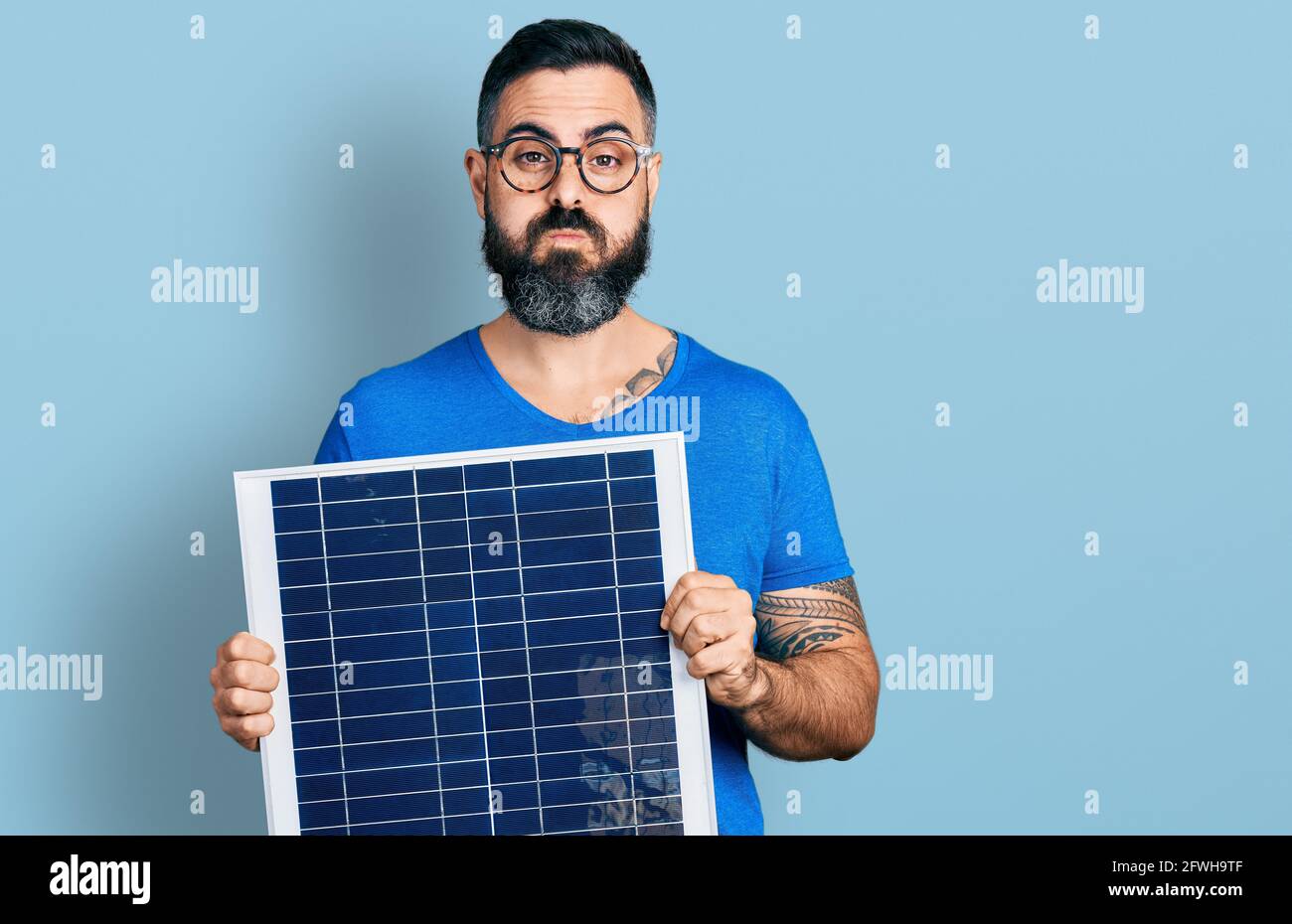 Hispanic man with beard holding photovoltaic solar panel puffing cheeks ...